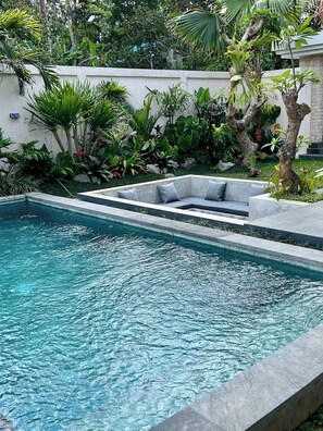 Piscina retangular com área de estar integrada e diversas plantas tropicais ao redor.