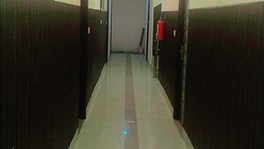 Um corredor comprido e estreito com piso refletivo, portas em ambos os lados, e um extintor de incêndio vermelho na parede direita.