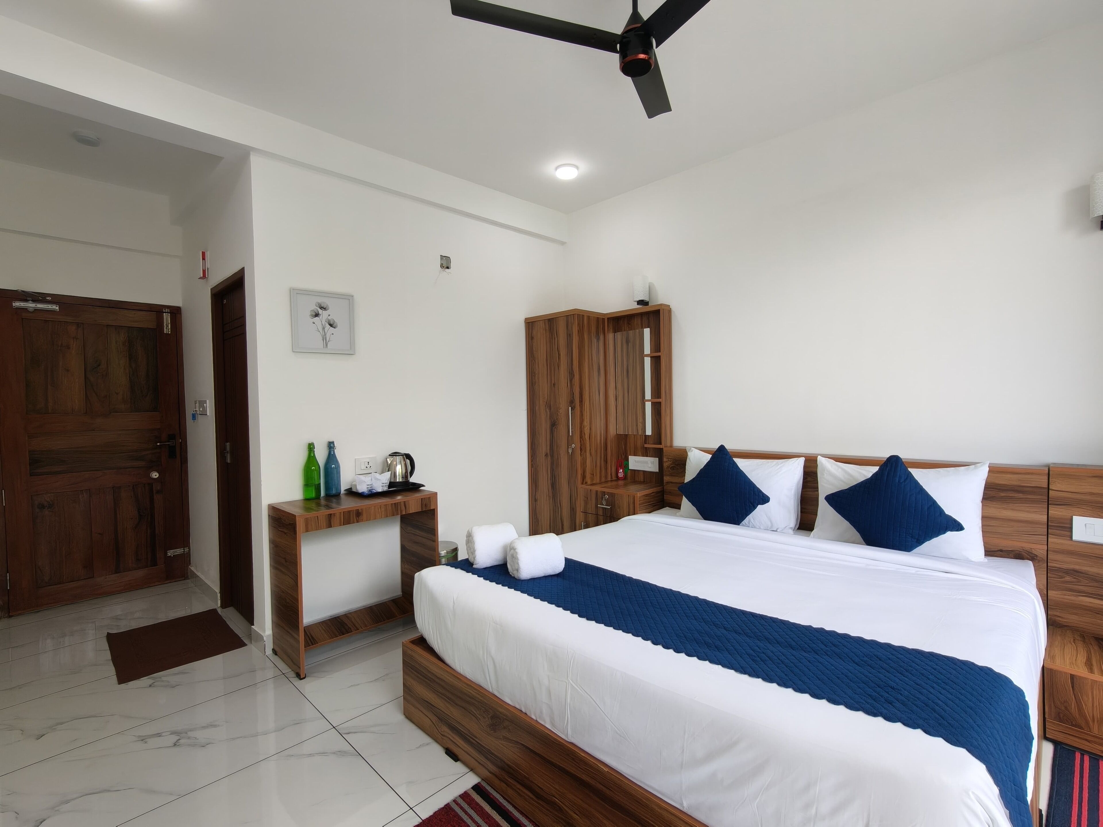 Quarto de hotel com uma cama grande, guarda-roupa de madeira, mesa de cabeceira com chaleira e ventoinha no teto.