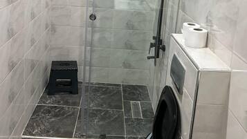 Box de chuveiro com paredes de vidro, chuveiro manual, prateleira pequena com artigos de higiene e vaso sanitário com assento preto.