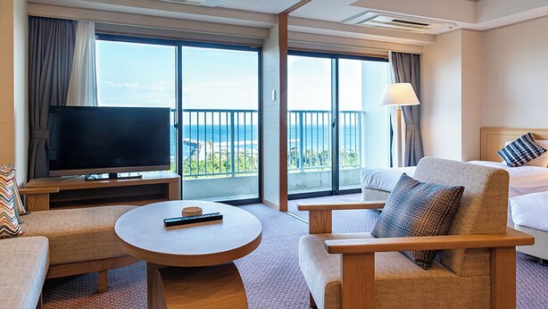 Quarto de hotel com uma televisão grande, um sofá, uma cadeira, uma mesa pequena e uma cama com vista para o oceano.