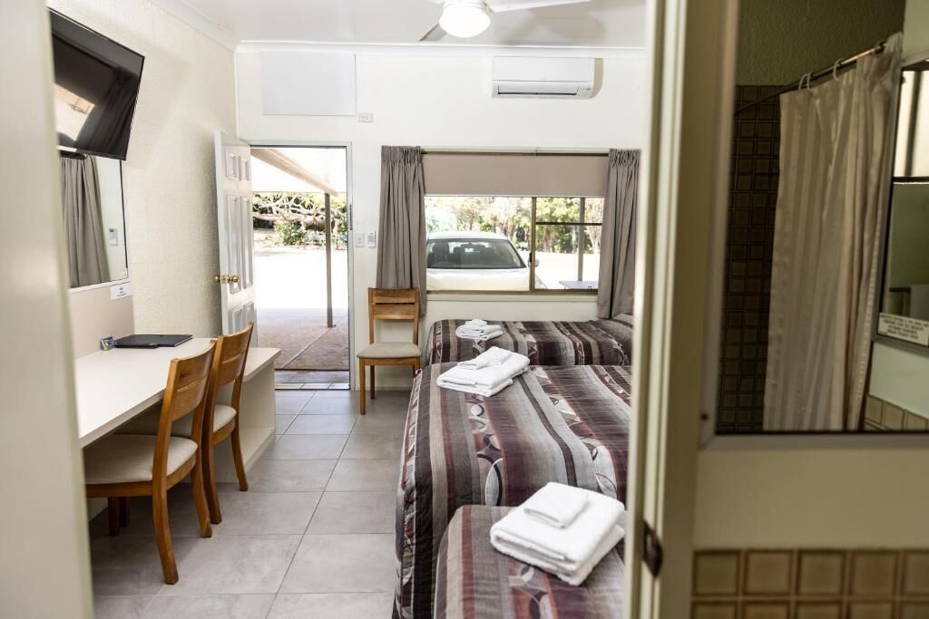 Quarto de hotel com uma cama, uma mesa e uma cadeira. Há uma janela com cortinas, e um carro é visível do lado de fora.