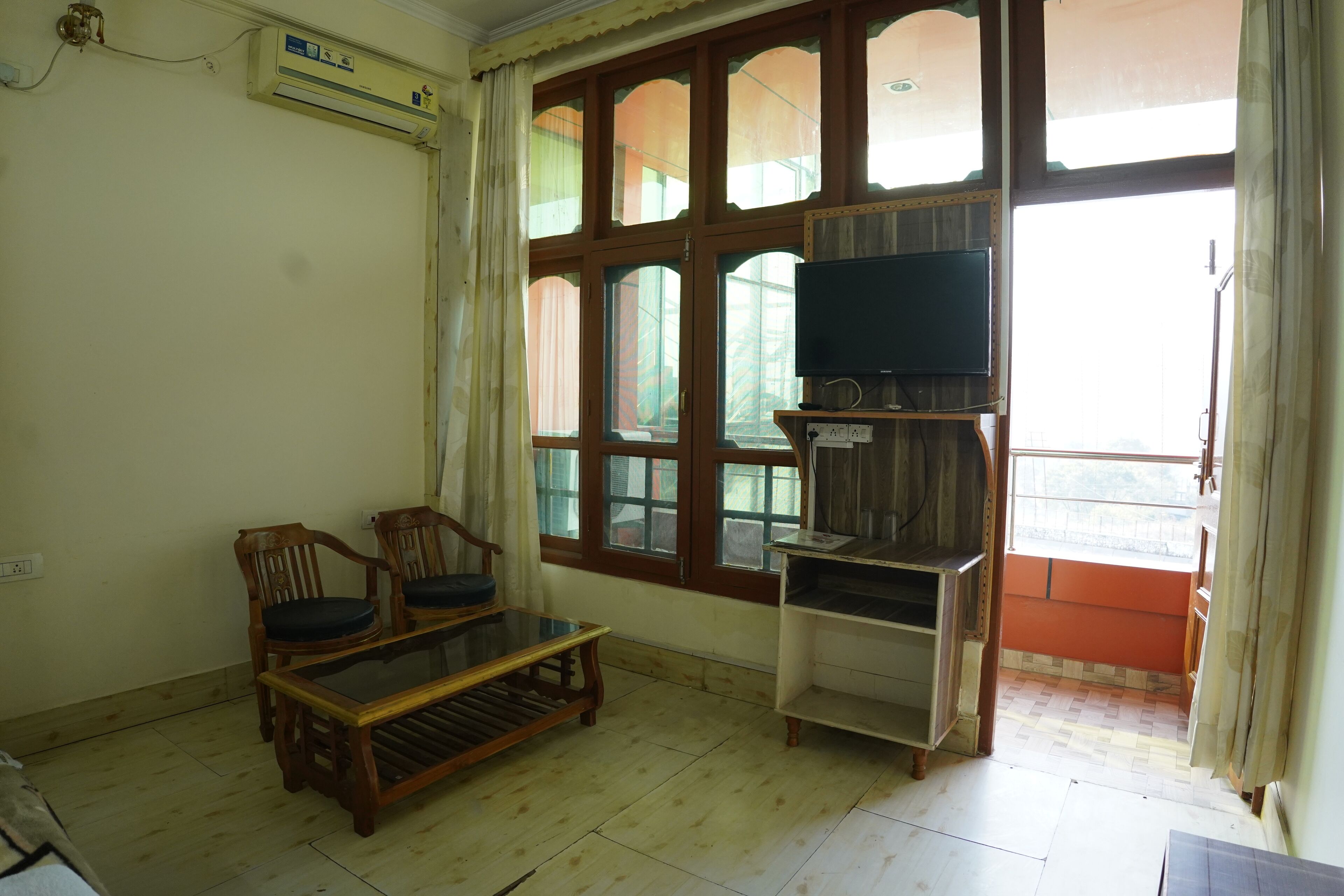 Um quarto com uma mesa e cadeiras de madeira, uma televisão sobre um móvel e janelas amplas com cortinas.