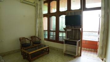 Um quarto com uma mesa e cadeiras de madeira, uma televisão sobre um móvel e janelas amplas com cortinas.