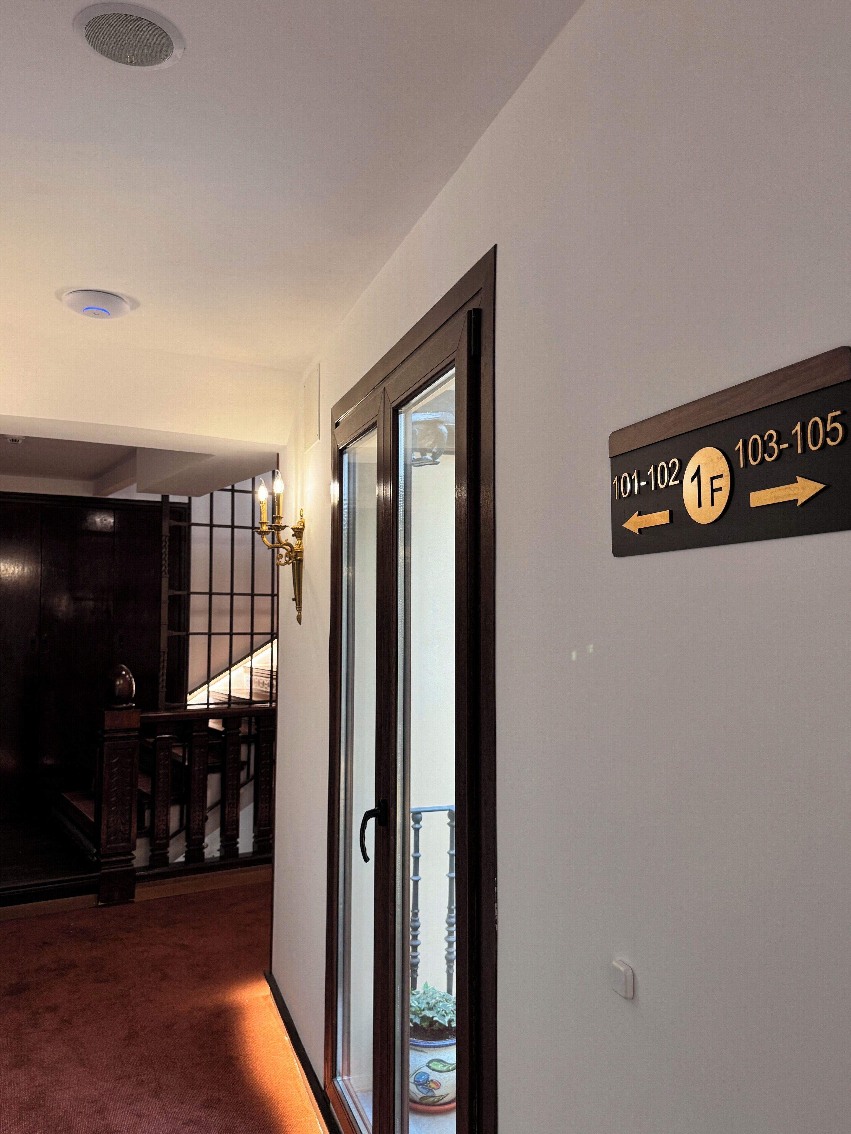 Corredor com placa indicando os quartos 101-102 e 103-105, escada e vista para uma varanda com plantas em vasos.