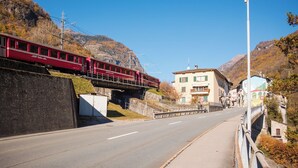 Um trem vermelho sobre uma ponte, acima de uma estrada, com edifícios e montanhas ao fundo.