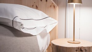Uma cama com cabeceira de madeira, uma mesa de cabeceira redonda também de madeira e um travesseiro branco com detalhes em costura preta.