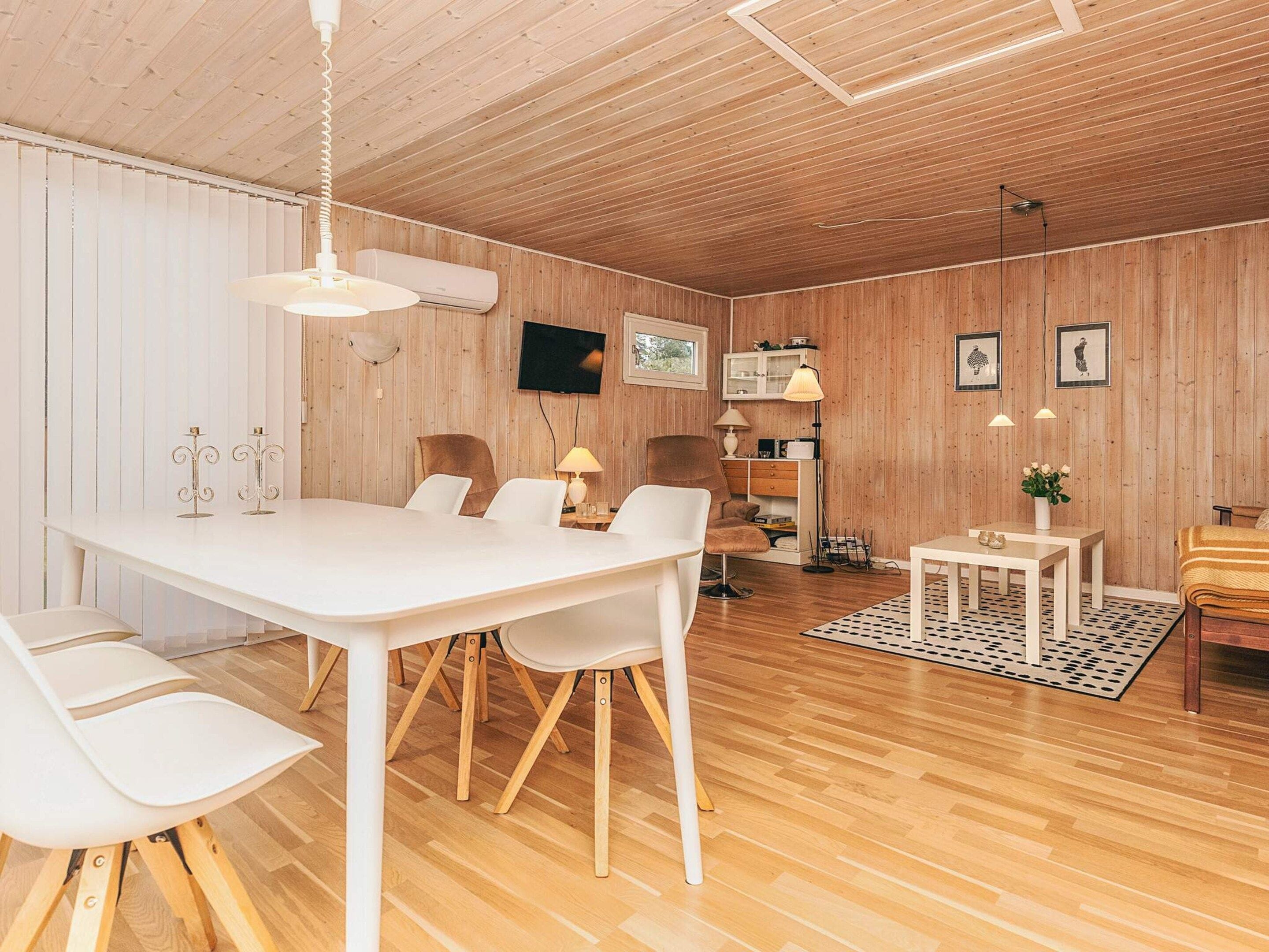 Uma sala de estar moderna com uma mesa de jantar branca, cadeiras e um sofá. Há uma TV fixada na parede, um abajur e uma pequena mesa lateral com um vaso.