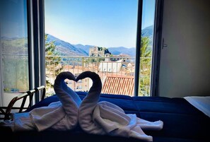 Uma cama com arranjo de toalhas em formato de coração, uma janela com vista para um edifício histórico e montanhas.
