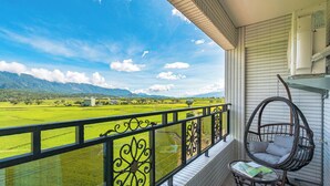 Uma varanda com vista para um campo verde e montanhas, uma cadeira suspensa e uma pequena mesa com uma revista.