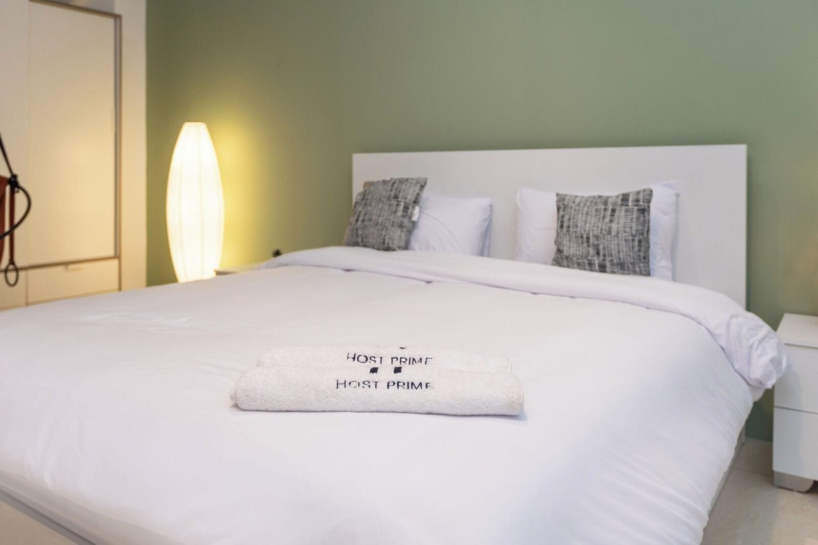 Cama bem arrumada com lençóis e almofadas brancos, luminária de cabeceira e toalha com a inscrição 'HOST PRIME'.