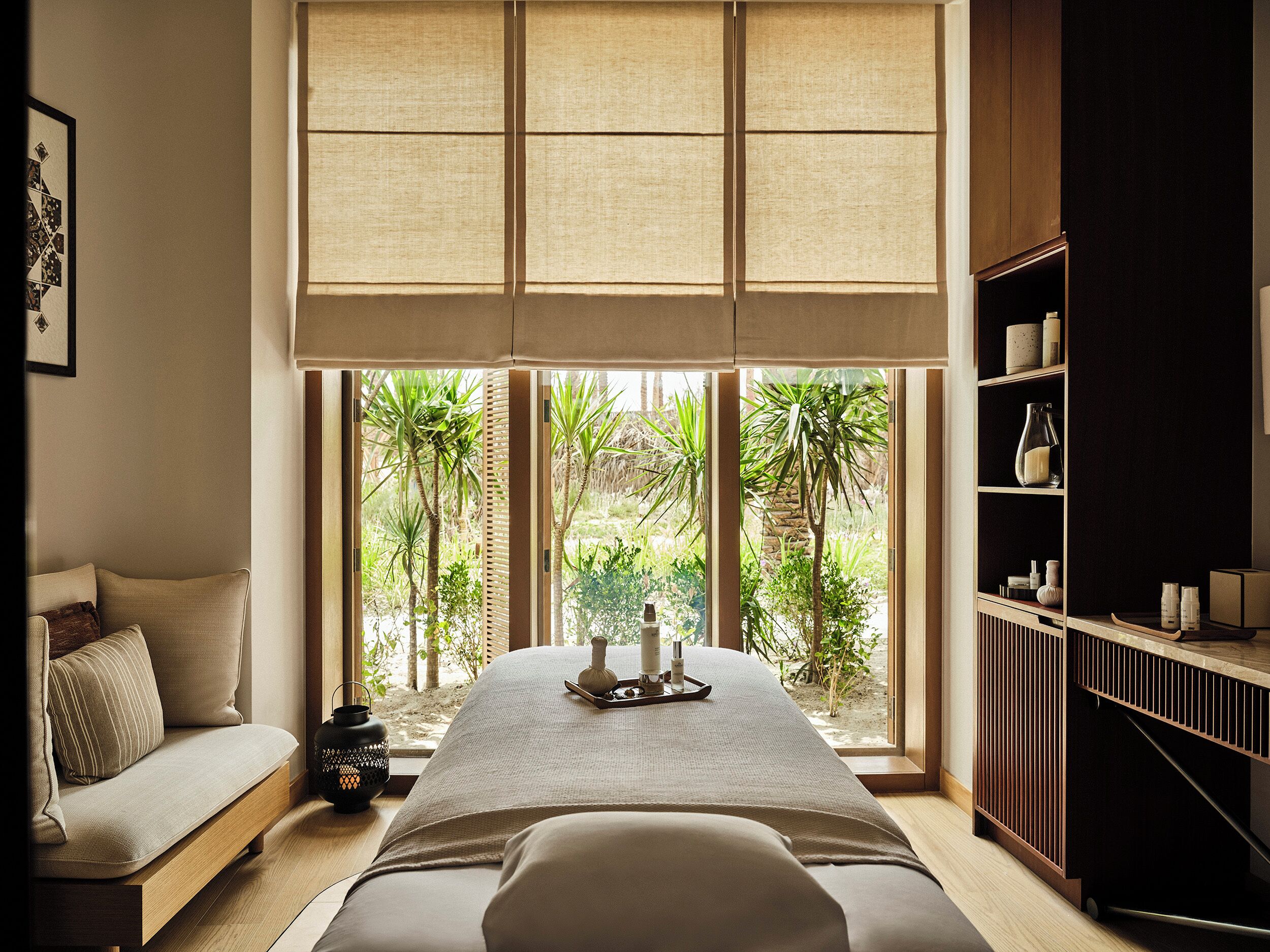 Um quarto de spa com mesa de massagem, um sofá, estante de livros e vista para o verde através de grandes janelas.