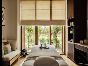 Um quarto de spa com mesa de massagem, um sofá, estante de livros e vista para o verde através de grandes janelas.