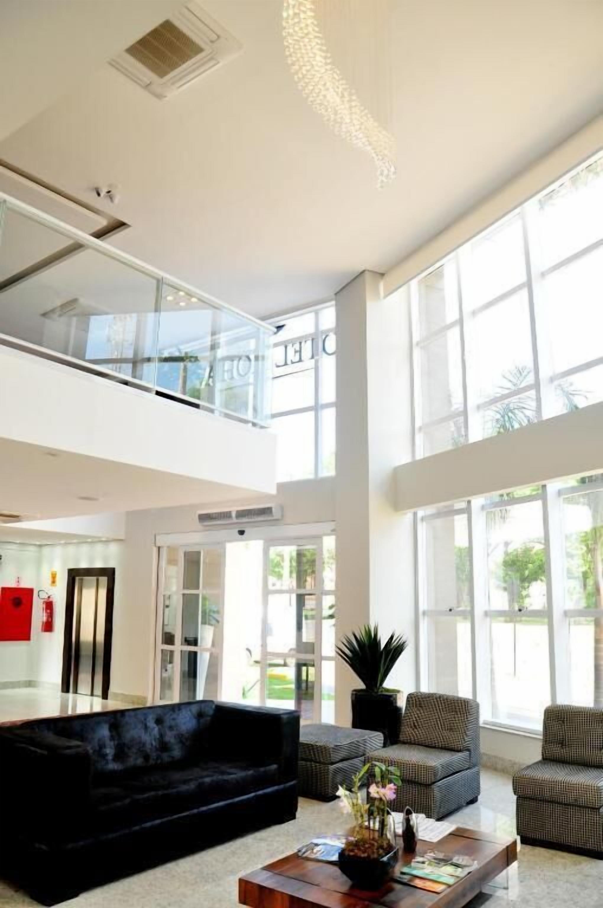 Um hall amplo com um sofá preto, poltronas estampadas e uma mesa de centro. Um guarda-corpo de vidro oferece vista para o nível inferior, e grandes janelas proporcionam luz natural.