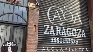 Vitrine fechada com uma grande persiana metálica, um letreiro com a inscrição 'CASA ZARAGOZA ALOJAMIENTOS' e um número de contato '3951151579'.