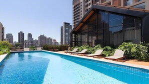 Piscina no terraço com espreguiçadeiras, rodeada por vegetação, e um horizonte urbano ao fundo.