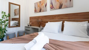 Quarto de hotel com uma cama grande, cabeceira de madeira, mesa de cabeceira com abajur, espelho e obra de arte abstrata na parede.