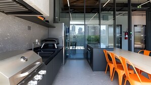 Uma cozinha moderna com churrasqueira, uma área de refeições com cadeiras laranjas e vista para a cidade através de grandes janelas.