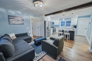 Uma sala de estar moderna com sofá seccional, banqueta e área de refeições, com cozinha ao fundo.