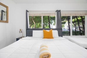 Quarto de hotel com uma cama grande, uma cama menor, uma janela com cortinas, uma mesa de cabeceira com abajur e um espelho na parede.