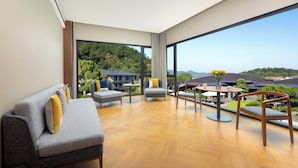 Uma sala de estar moderna com um sofá cinza, duas poltronas, uma mesa de jantar de madeira e uma grande janela com vista para uma paisagem verde exuberante e edifícios distantes.