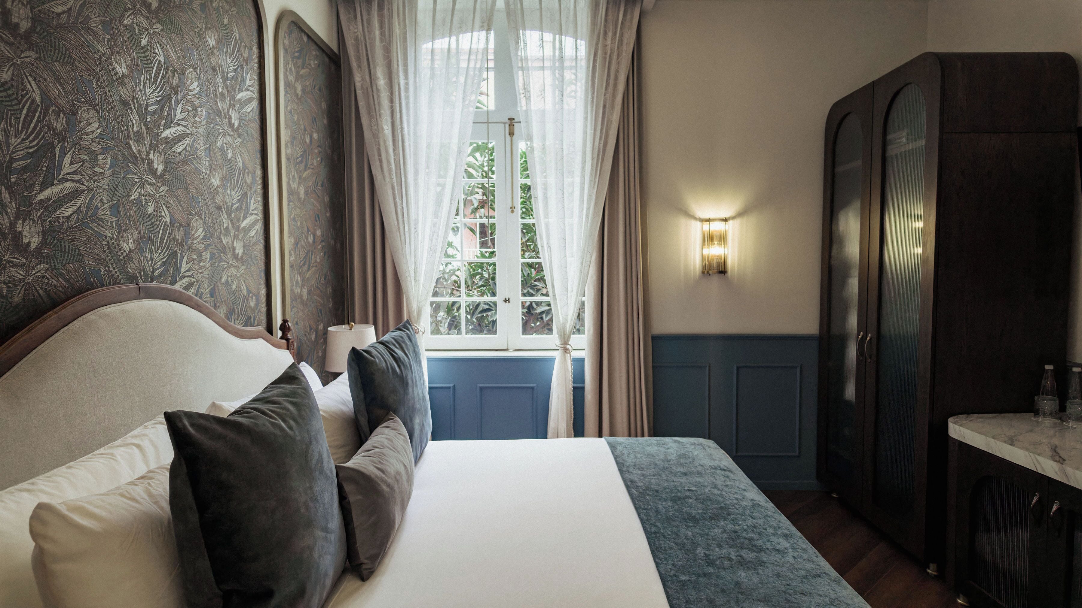 Quarto de hotel com cama, guarda-roupa, janela com cortinas e luminária na parede.