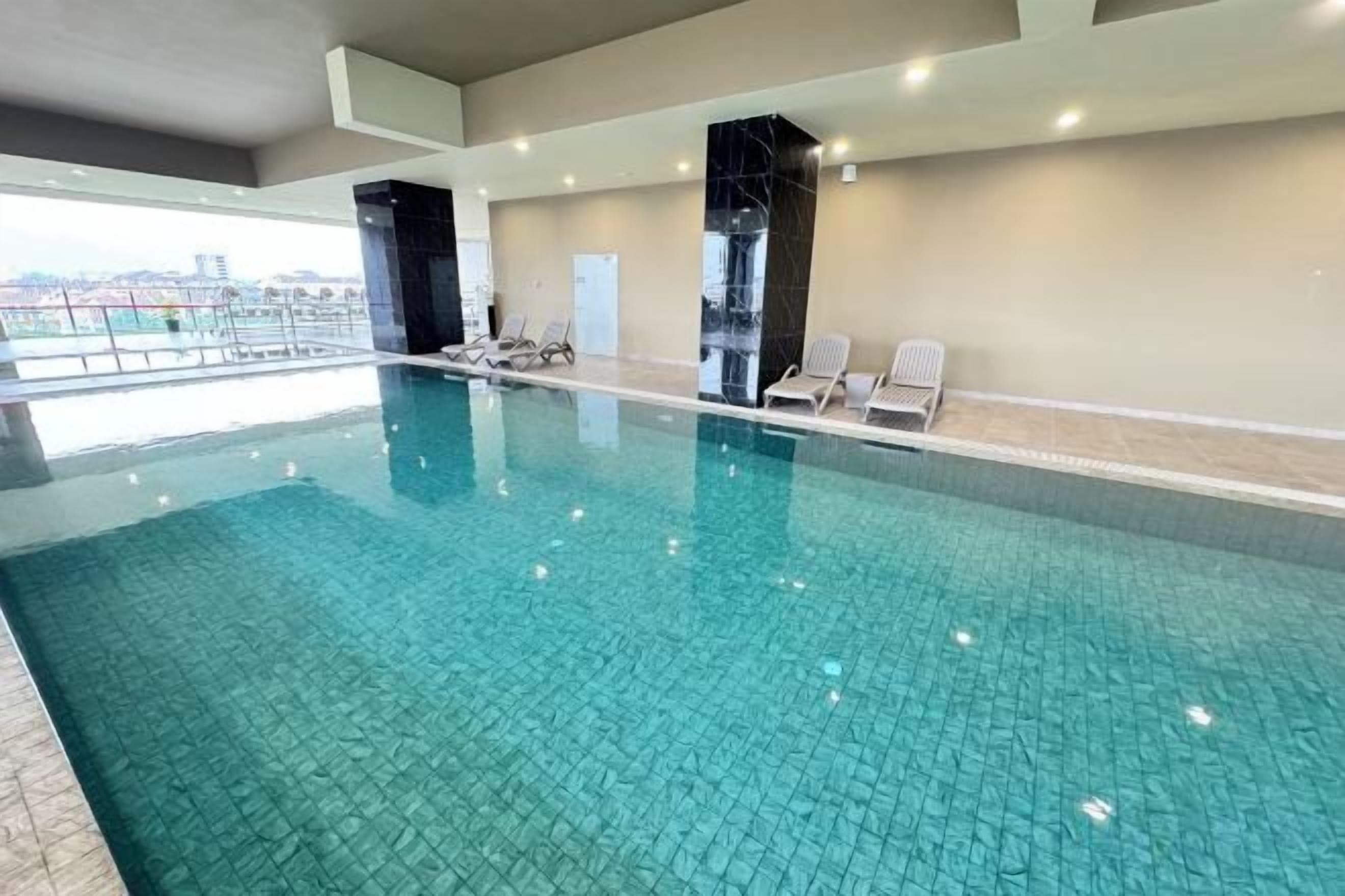 Piscina interna com água cristalina, rodeada por espreguiçadeiras e teto alto.