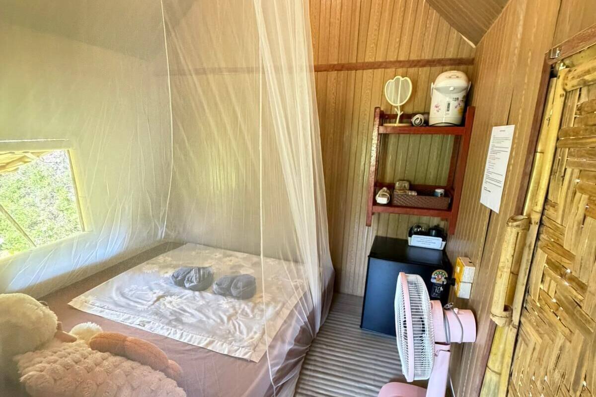 Quarto com cama, ventilador ao lado, prateleira pequena com produtos de higiene pessoal e janela com vista.