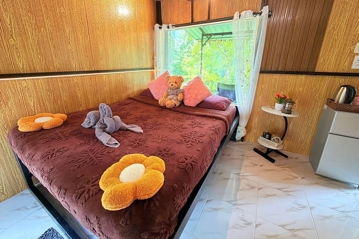 Quarto com cama, travesseiros, um urso de pelúcia, uma mesinha com chaleira e vista para o verde lá fora.