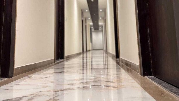 Um corredor comprido e estreito, com chão de mármore polido e portas de madeira escura em ambos os lados.