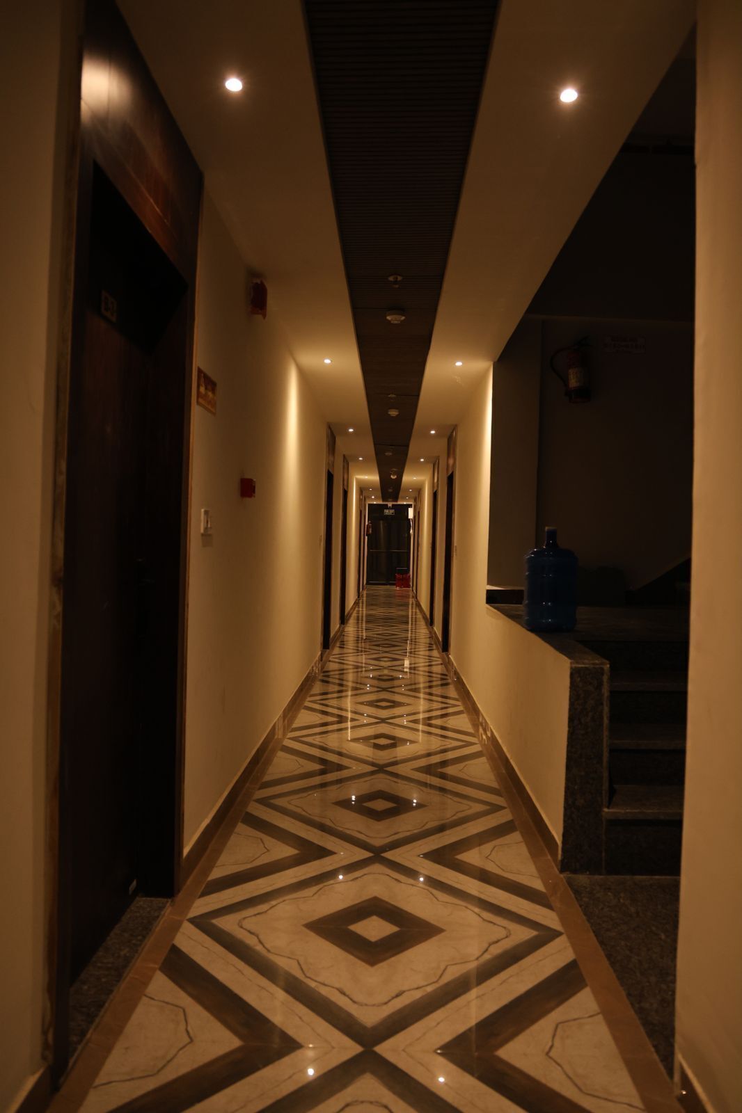 Um corredor de hotel com piso estampado em padrão geométrico, iluminação embutida e uma escada à direita.