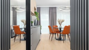 Uma área de refeições moderna com uma mesa central, cadeiras laranja e uma cozinha ao fundo.