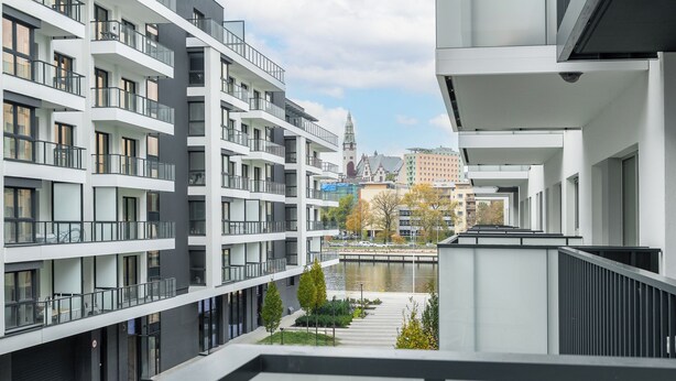 Um edifício residencial moderno de vários andares com varandas, um pátio interno e vista para um rio e edifícios históricos ao fundo.