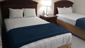 Quarto de hotel com duas camas, cada uma com cabeceira de madeira, roupa de cama branca e um cobertor azul em uma das camas. Há uma mesa de cabeceira com uma luminária e um telefone.