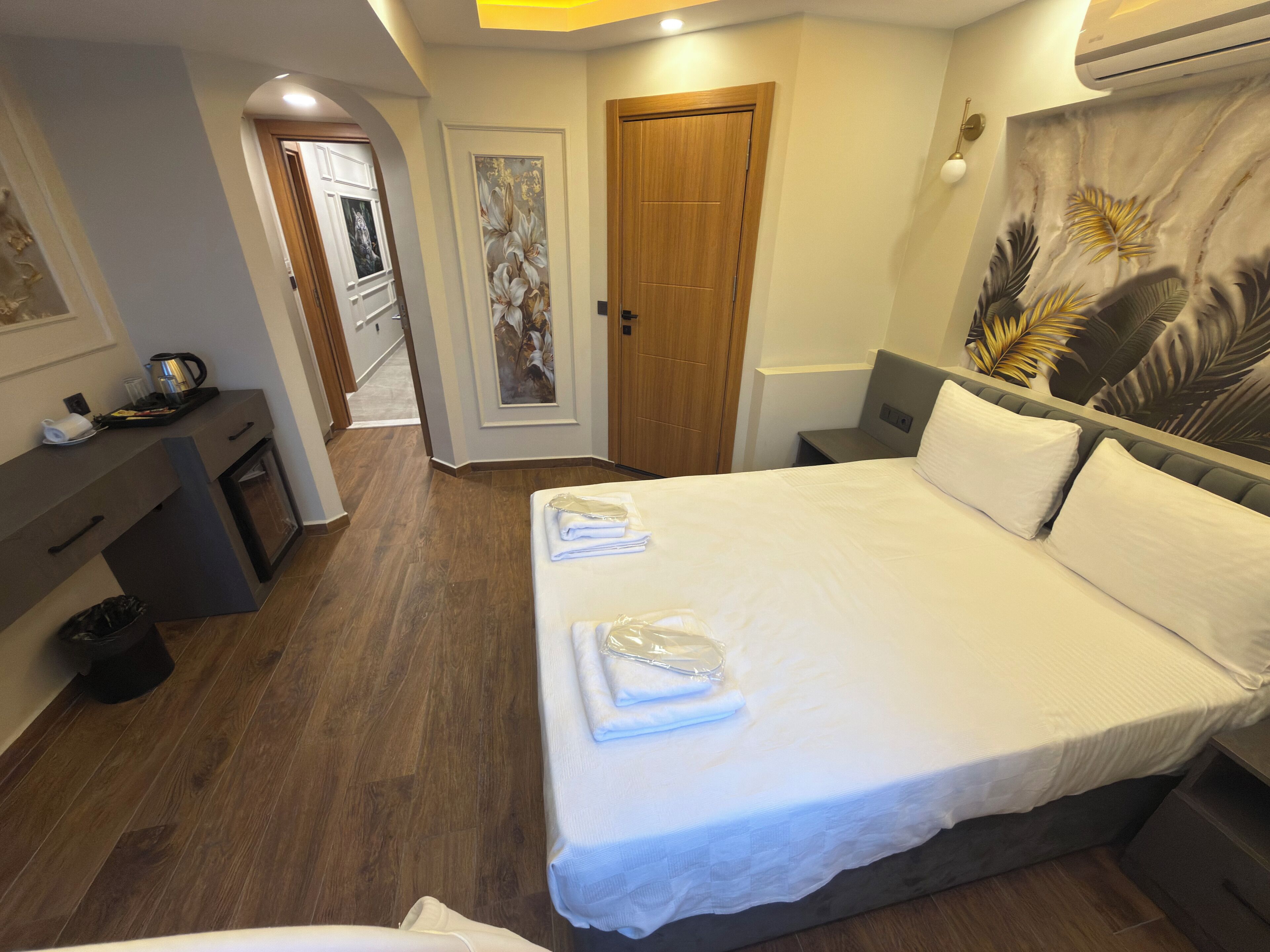 Quarto de hotel com uma cama, uma pequena escrivaninha, uma cadeira, um cesto de lixo e uma porta.