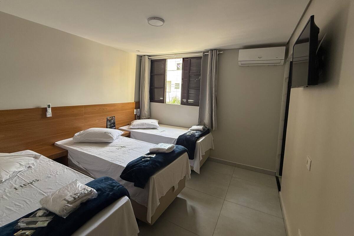 Quarto de hotel com duas camas, cabeceira de madeira, janela com cortinas, ar condicionado e televisão de tela plana na parede.