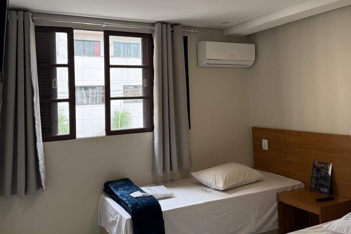 Quarto de hotel com duas camas, uma janela com cortinas, ar condicionado e cabeceira de madeira.