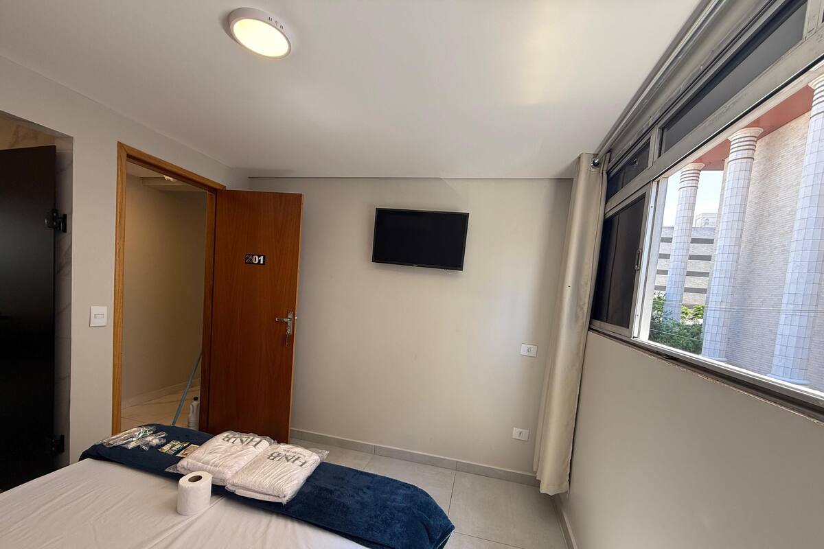 Quarto de hotel com cama, televisão na parede, porta com número e janela com vista para prédios.