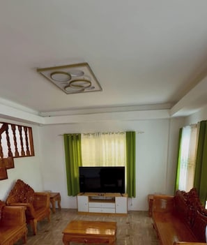 Uma sala de estar com televisão sobre um suporte de madeira, cortinas verdes e mobiliário de madeira.