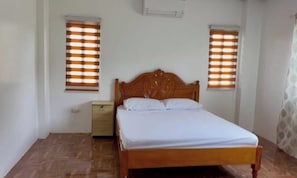 Quarto com cama de madeira, duas janelas com persianas, uma pequena mesa de cabeceira e um ar condicionado fixado na parede.