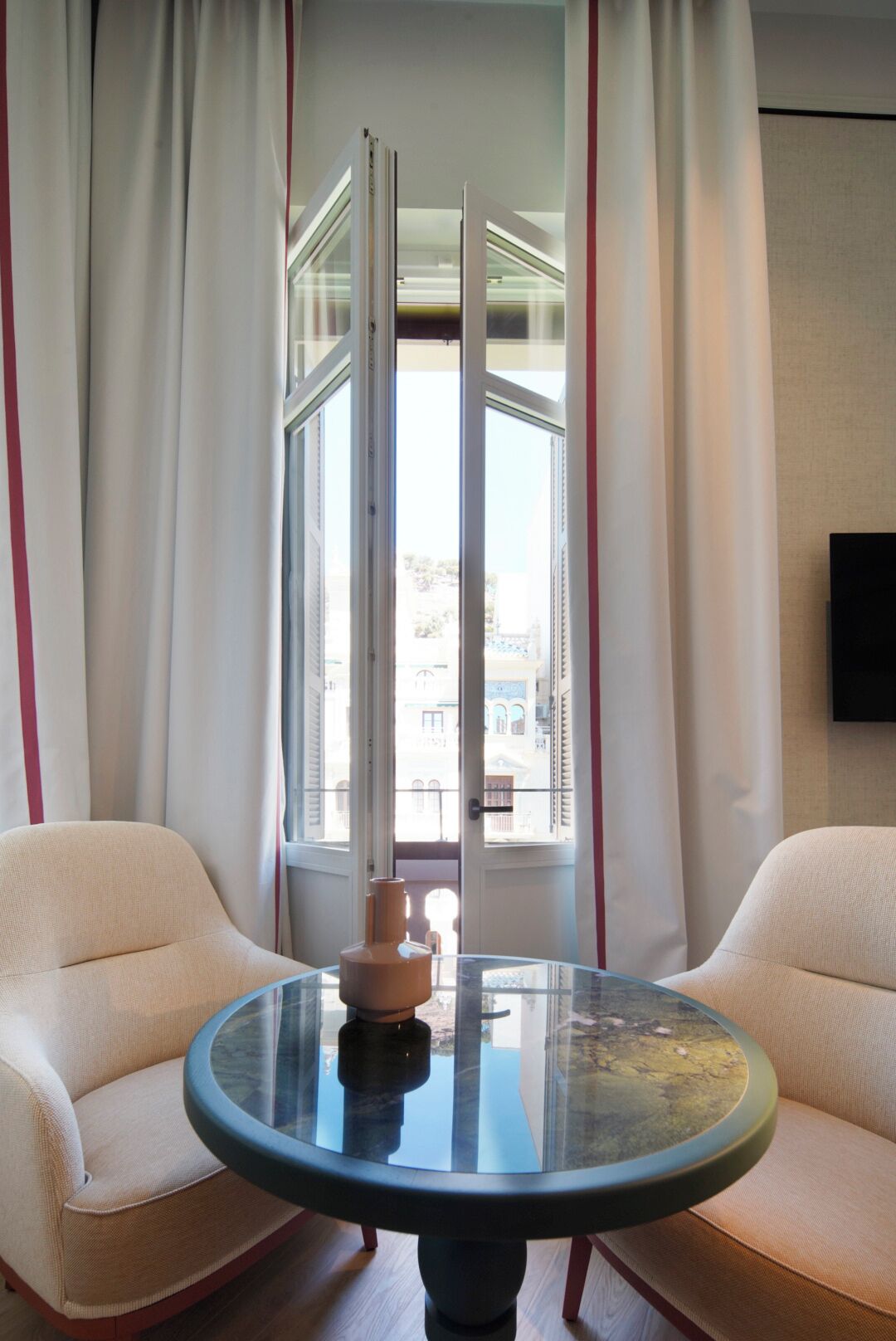 Quarto com mesa de vidro, duas cadeiras bege, um vaso e uma janela com cortinas.