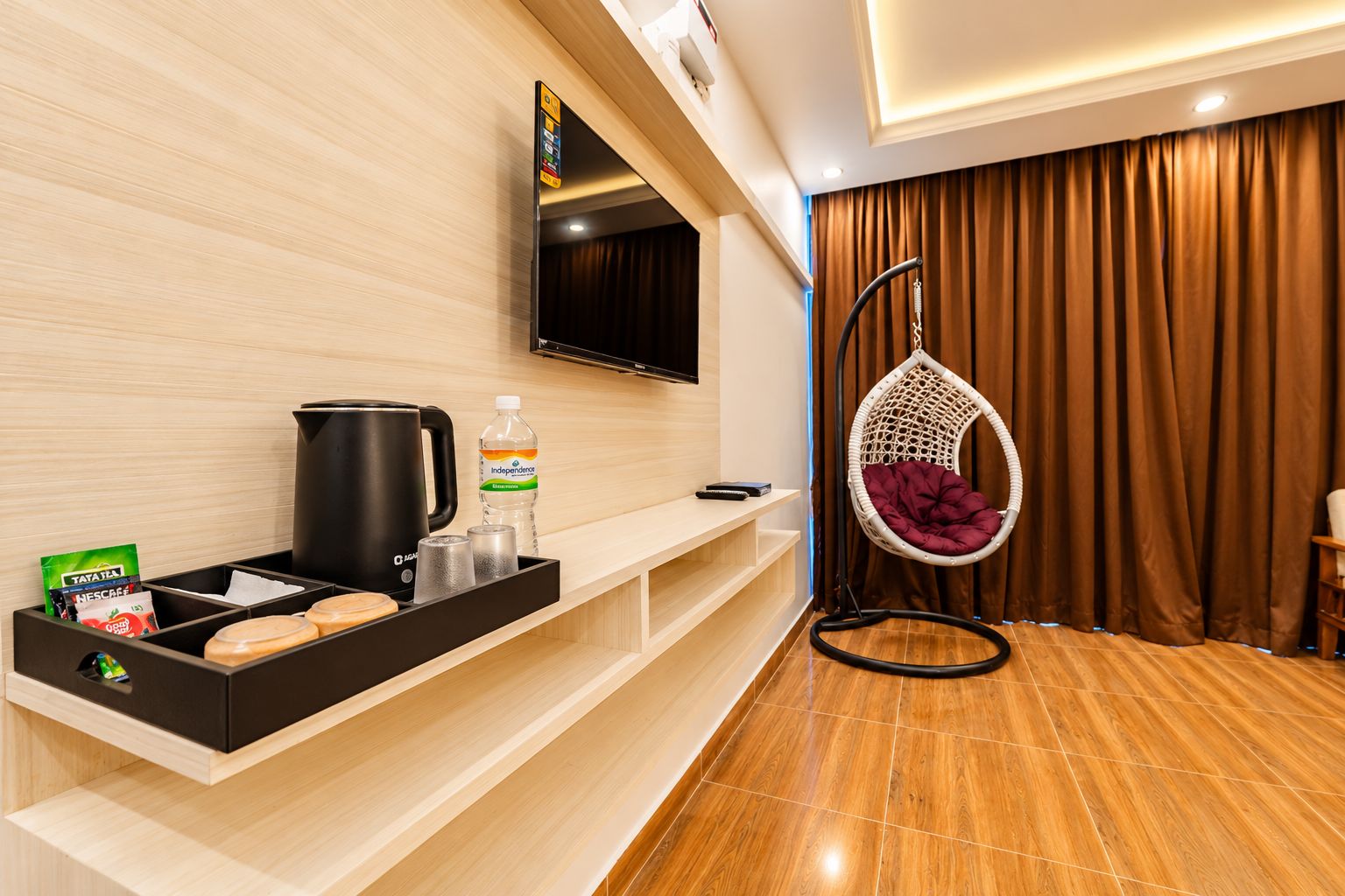 Quarto de hotel com piso de madeira, cafeteira preta, garrafa de água, cadeira suspensa e televisão de tela plana fixada na parede.