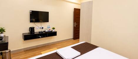 Quarto de hotel com cama, televisão na parede, pequeno móvel com objetos e porta.