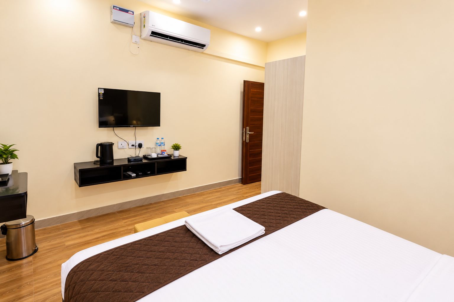 Quarto de hotel com cama, televisão na parede, pequeno móvel com objetos e porta.