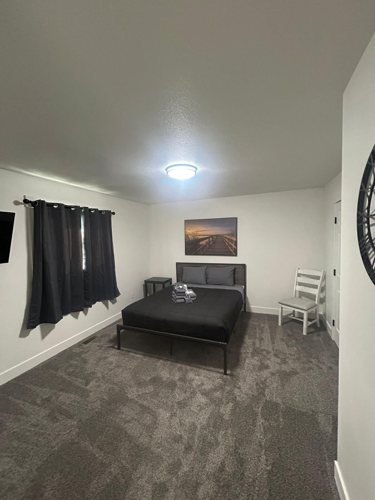 Quarto com cama, uma cadeira, uma mesinha, um quadro e janela com cortinas.