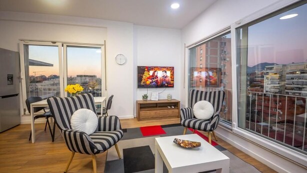 Uma sala de estar moderna com uma televisão grande, poltronas listradas, área de jantar e vista para a cidade.
