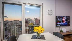 Quarto com uma janela grande, uma mesa branca com um vaso de flores amarelas, um relógio e uma televisão exibindo 'Being Eddie'.
