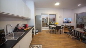 Cozinha moderna com pia, fogão e geladeira. Há uma área de refeições com mesa e cadeiras, e uma TV fixada na parede.