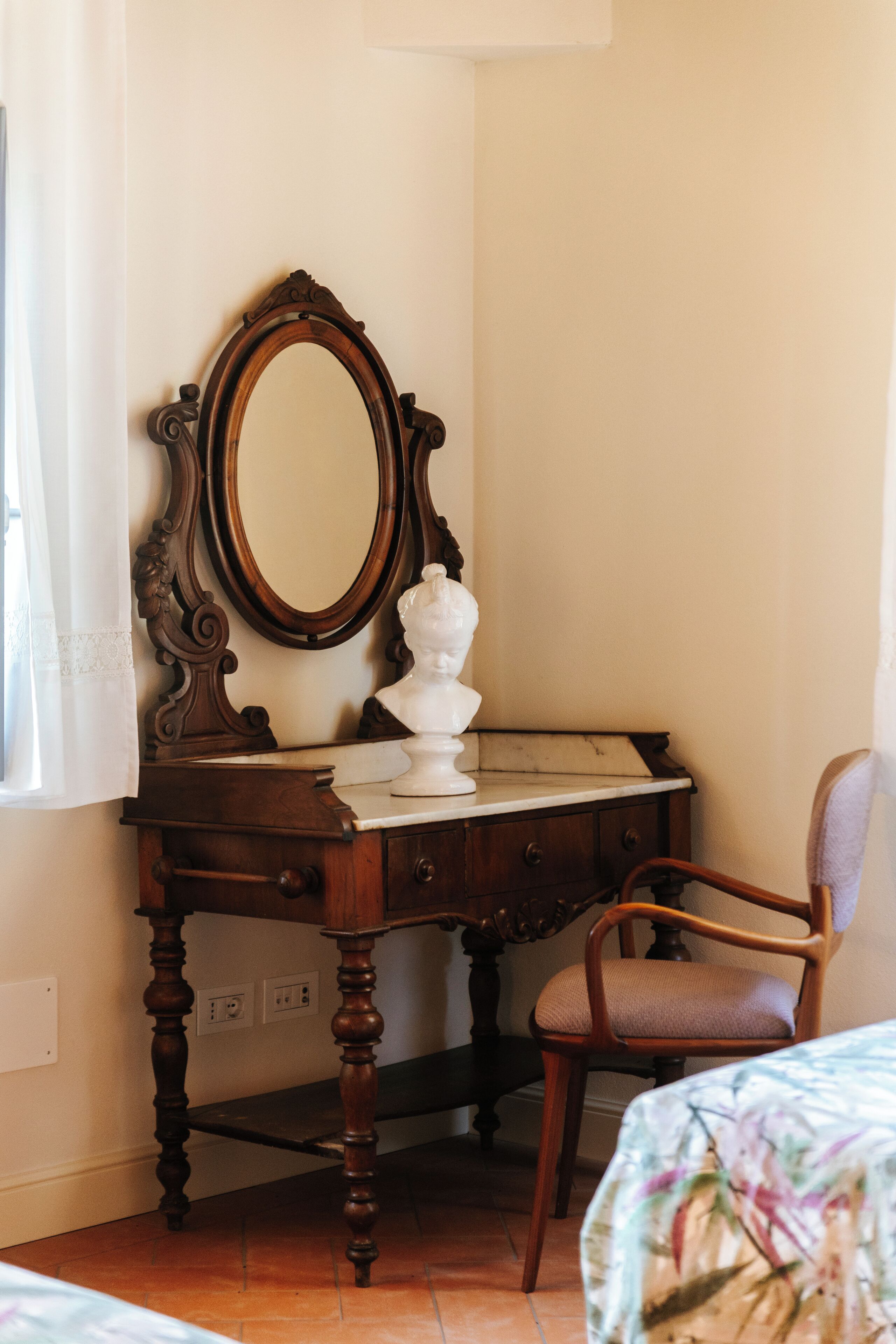 Quarto nos cantos com escrivaninha de madeira, uma cadeira, um espelho e um busto sobre a escrivaninha.