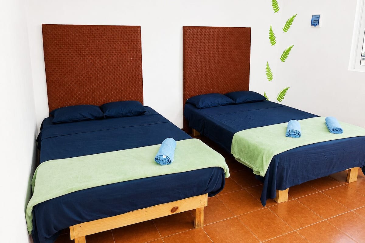 Duas camas de solteiro com estrutura de madeira, roupa de cama azul escura e toalhas verdes.